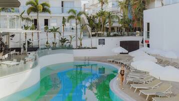 Piscina com espreguiçadeiras e guarda-sóis, cercada por palmeiras e um edifício branco.