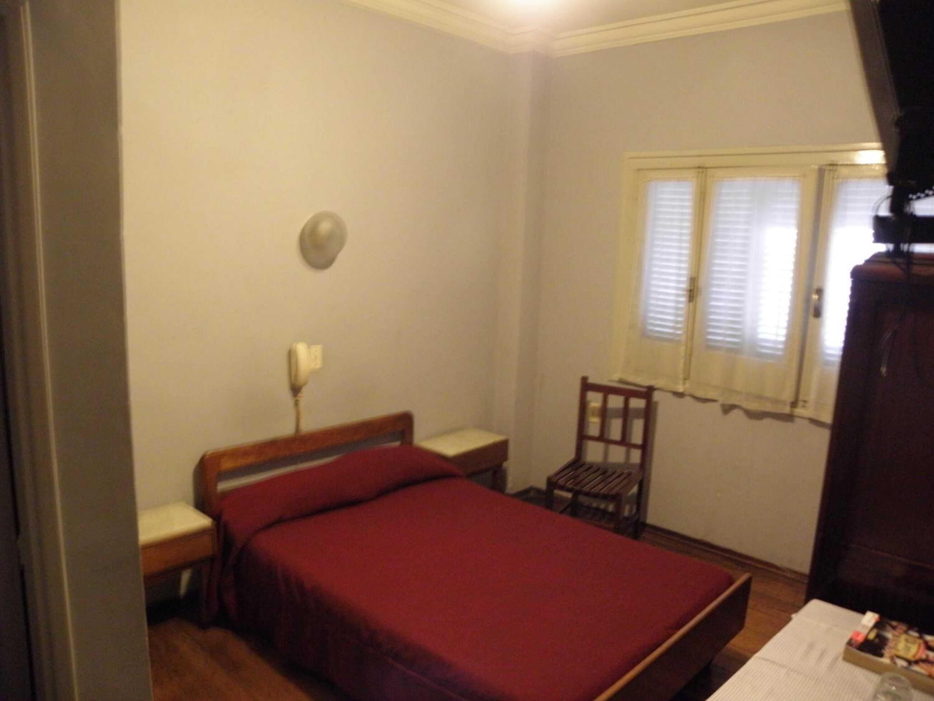 Quarto pequeno e simples com uma cama de solteiro, mesa de cabeceira, uma cadeira e uma janela com persianas.
