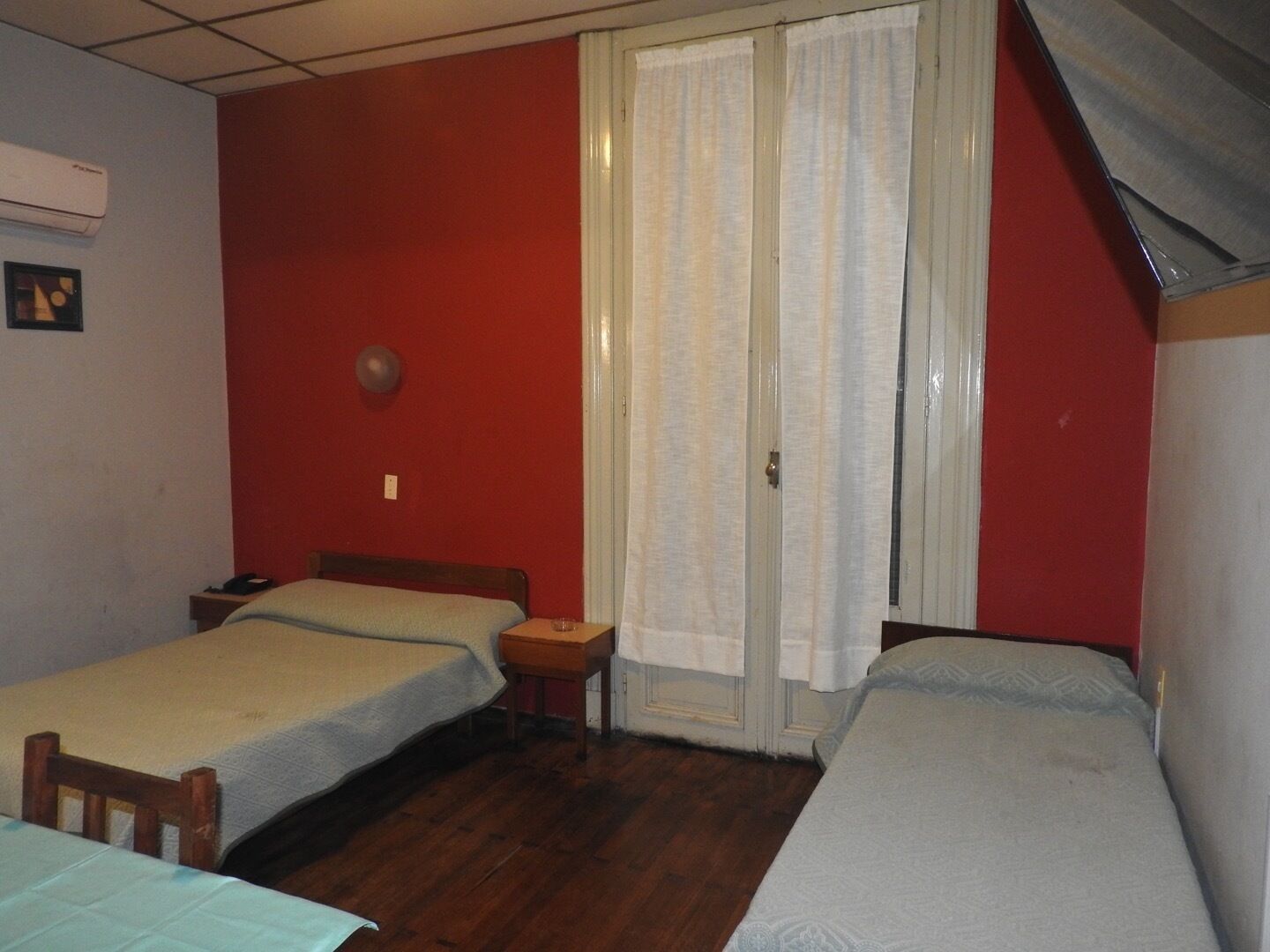 Quarto com duas camas de solteiro, uma mesinha, uma luminária na parede e janela com cortinas.