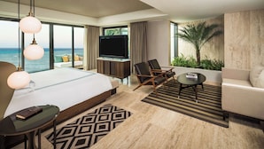 Quarto de hotel com uma cama grande, televisão, sofá, mesa pequena e vista para o oceano.