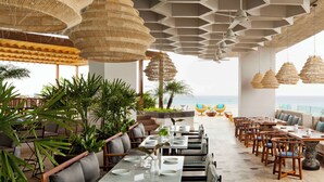 Um restaurante com vista para o oceano, com luminárias de vime suspensas, plantas verdes e mobiliário de madeira.