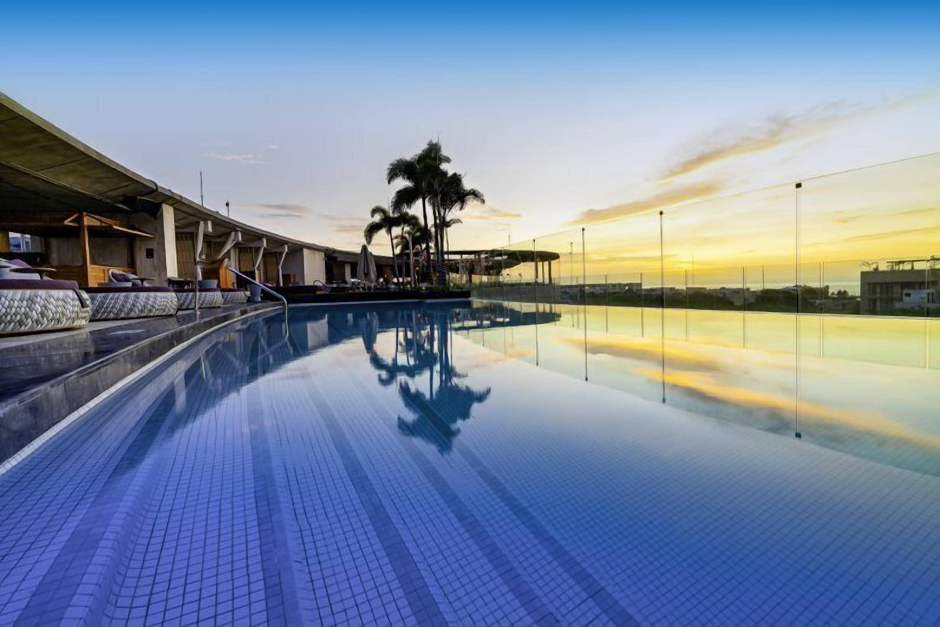 Uma piscina moderna infinita com vista desimpedida para o pôr do sol, palmeiras e uma área de estar coberta.