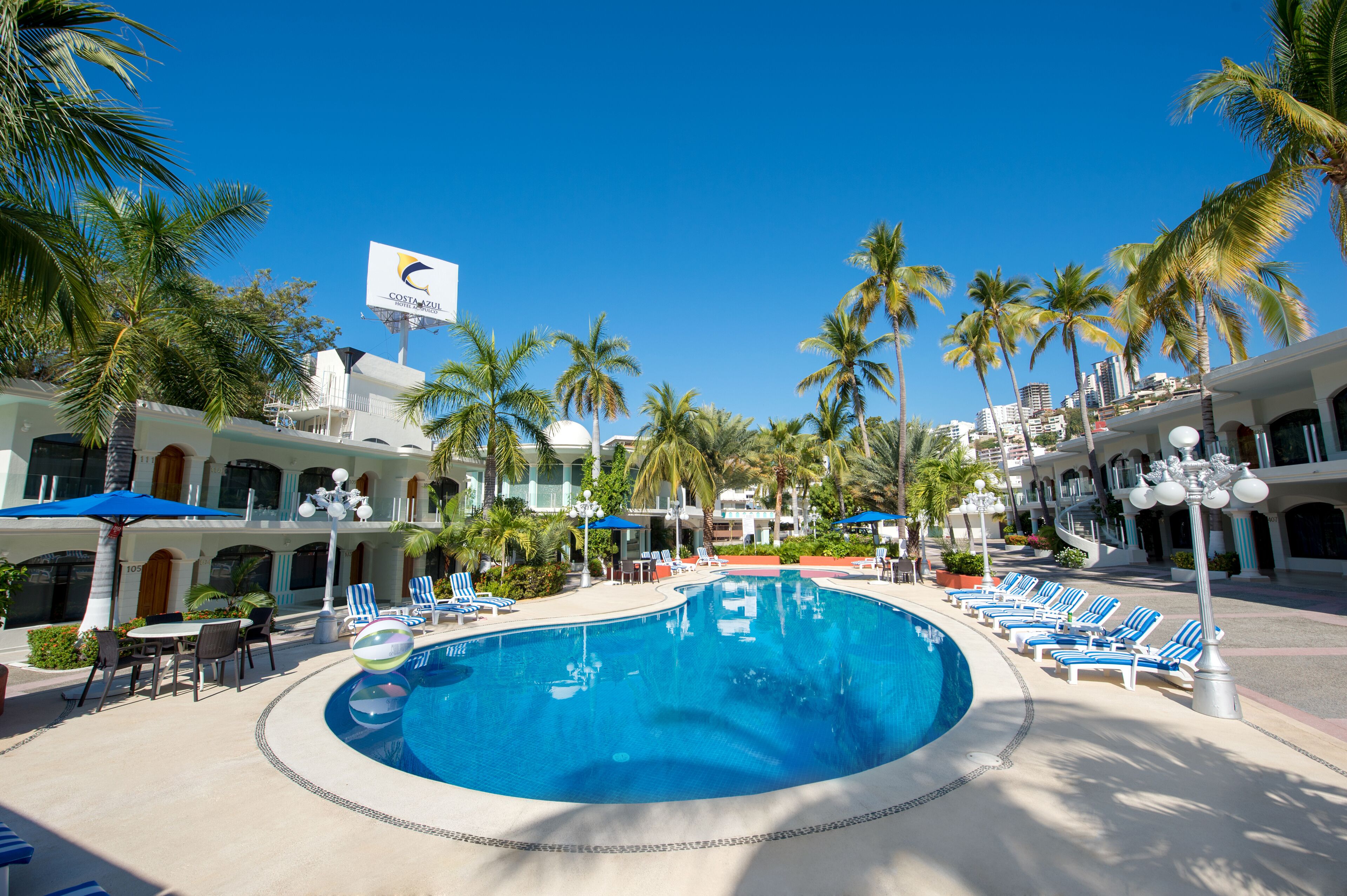 Área de piscina de resort, com palmeiras, espreguiçadeiras e um prédio grande com uma placa.