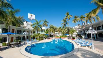 Área de piscina de resort, com palmeiras, espreguiçadeiras e um prédio grande com uma placa.