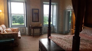 Quarto com cama de dossel, sofá, mesa pequena e janela com vista para o verde.