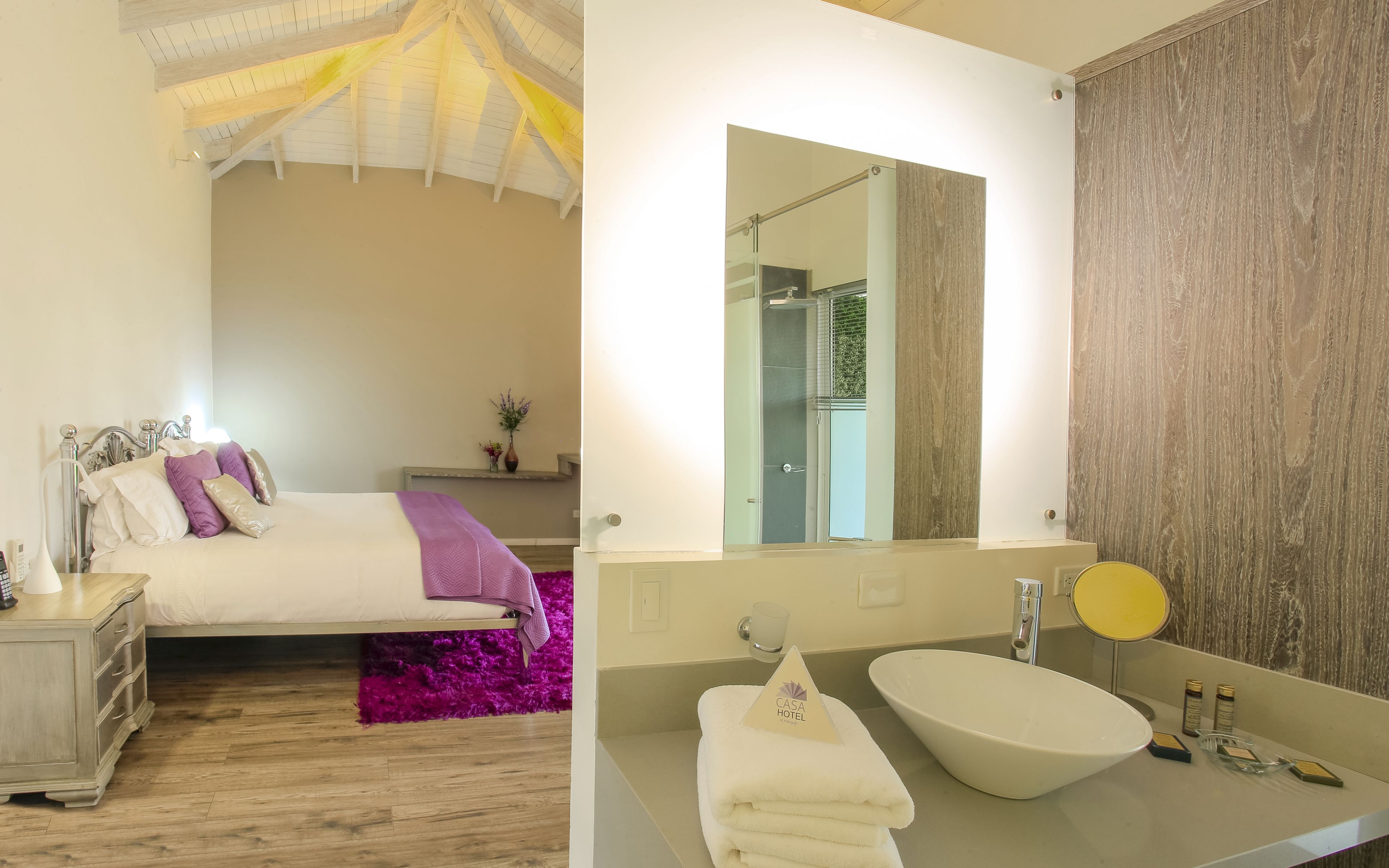 Quarto com cama, mesa de cabeceira e apoio para objetos pessoais. Inclui banheiro com pia, espelho e toalha.