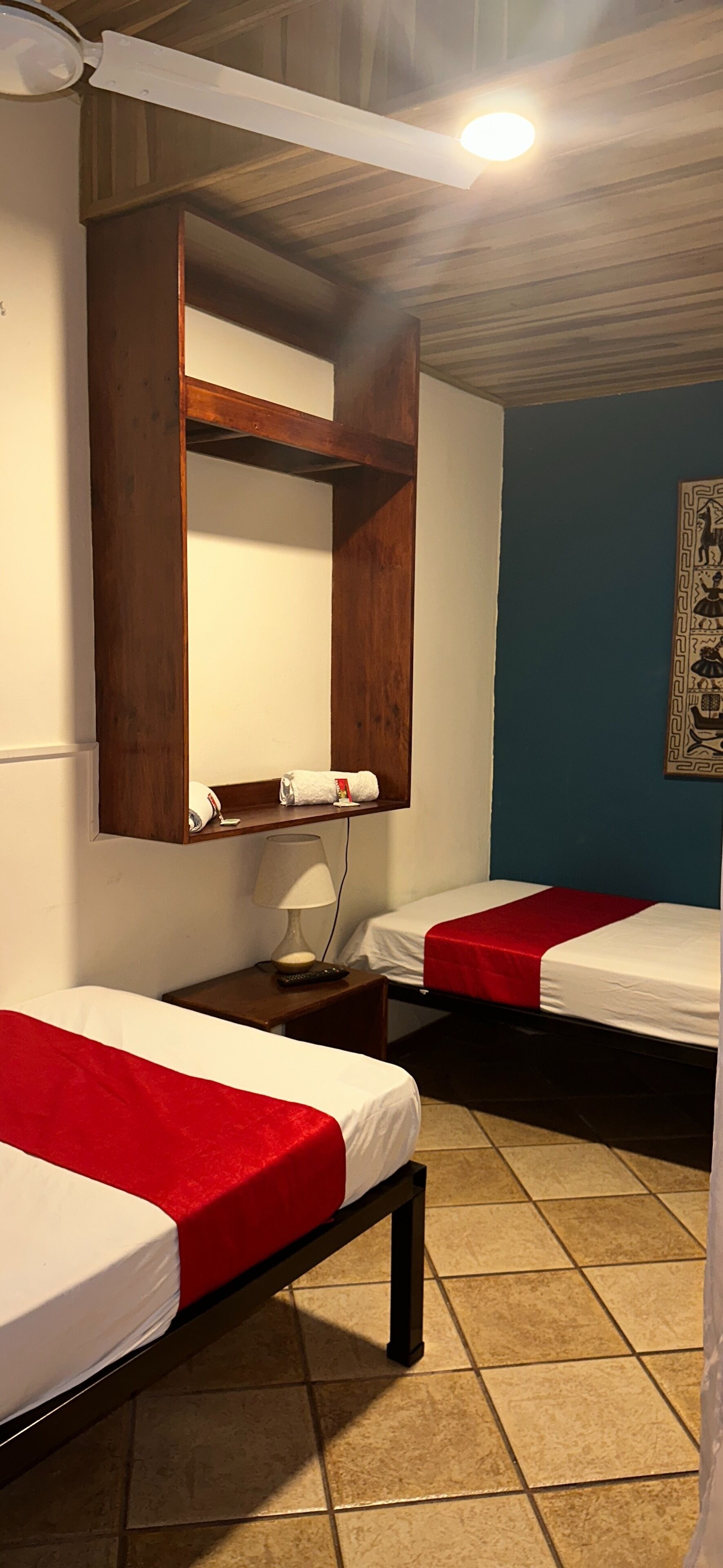 Quarto com duas camas, cada uma com colcha vermelha e branca. Uma prateleira de madeira sobre uma das camas acomoda um telefone e um abajur. O chão é de azulejos e a parede é azul, com um painel decorativo.