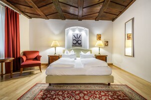 Quarto de hotel com uma cama grande, duas mesas de cabeceira com luminárias, uma poltrona vermelha, teto de madeira e uma grande obra de arte na parede.