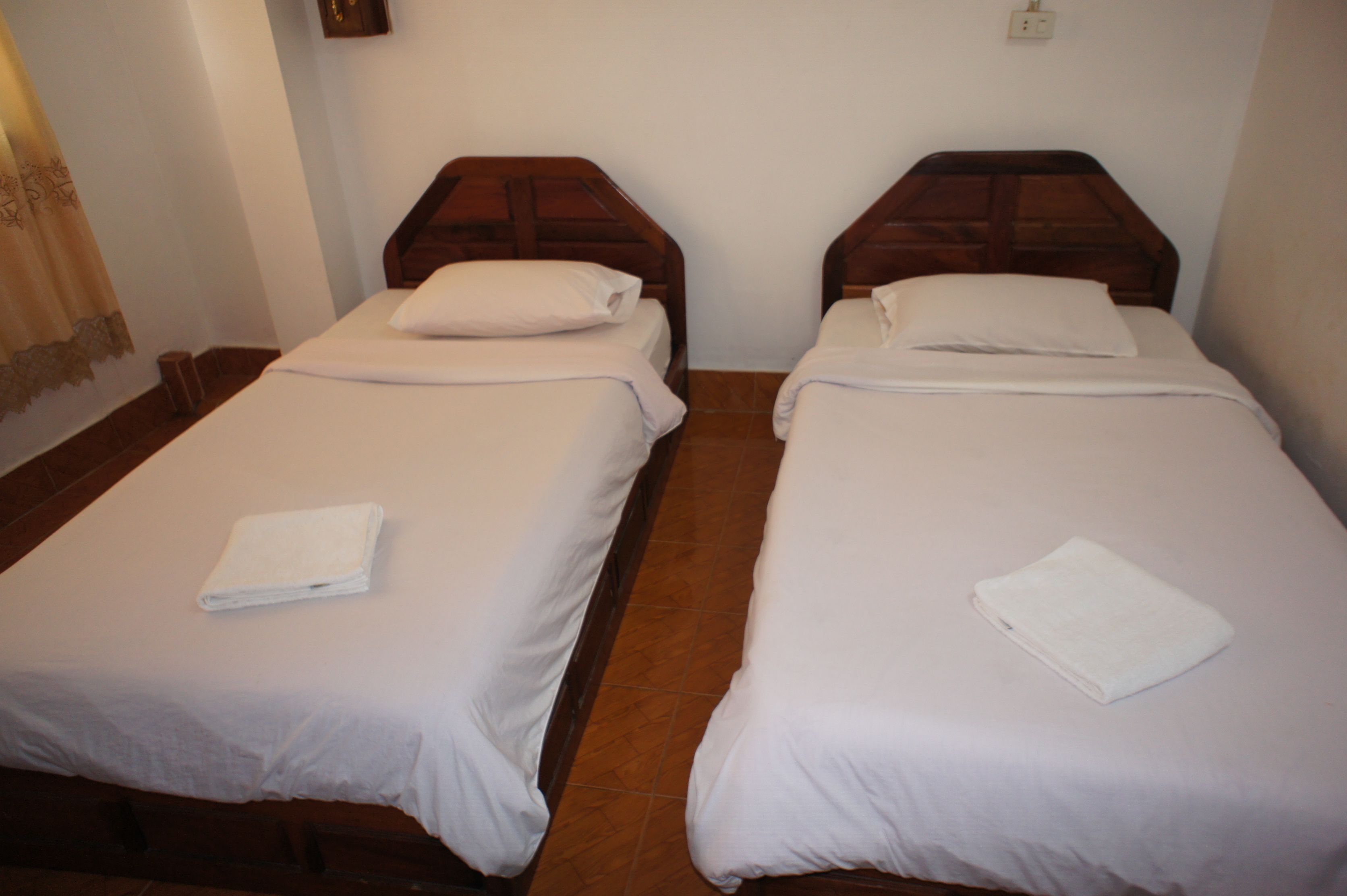 Duas camas de solteiro com cabeceiras de madeira e roupa de cama branca, incluindo toalhas, num quarto com chão em mosaico e uma tomada na parede.