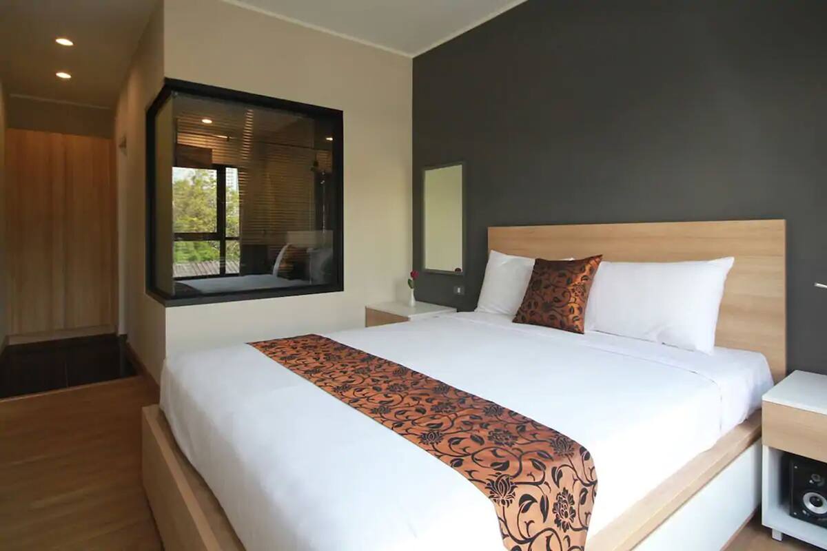 Quarto de hotel moderno com uma cama grande, janela com persianas, mesa de cabeceira e criado-mudo.