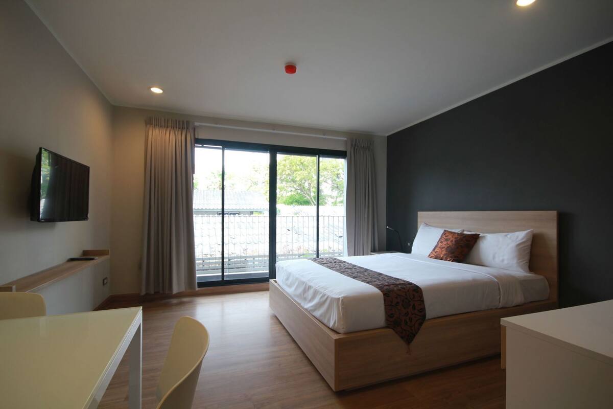Quarto de hotel com cama, televisão, escrivaninha e uma janela grande com cortinas.
