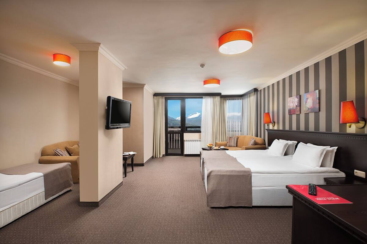 Quarto de hotel com cama, sofá, televisão, escrivaninha e janela com vista para montanhas.