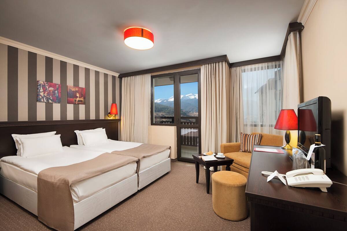 Quarto de hotel com uma cama, escrivaninha, cadeira, mesa, abajur, telefone e vista para as montanhas.