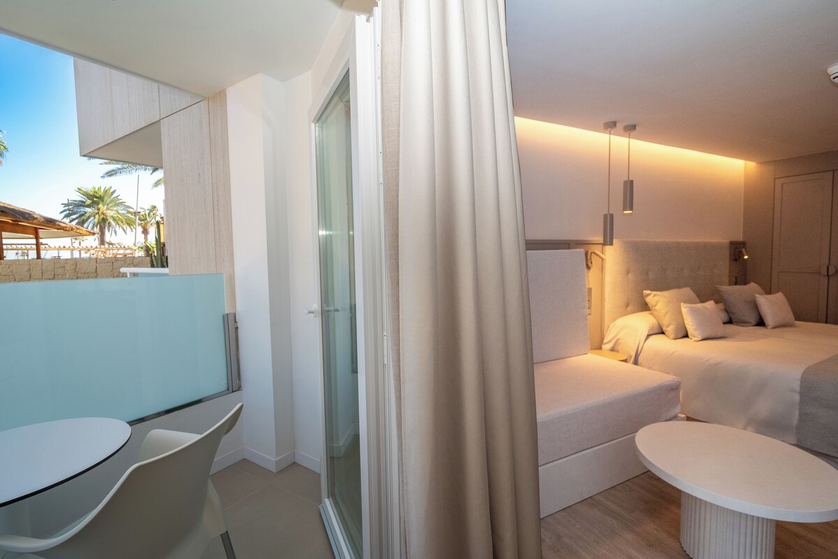 Quarto de hotel com cama, uma mesinha, uma cadeira, porta de vidro e vista para palmeiras.