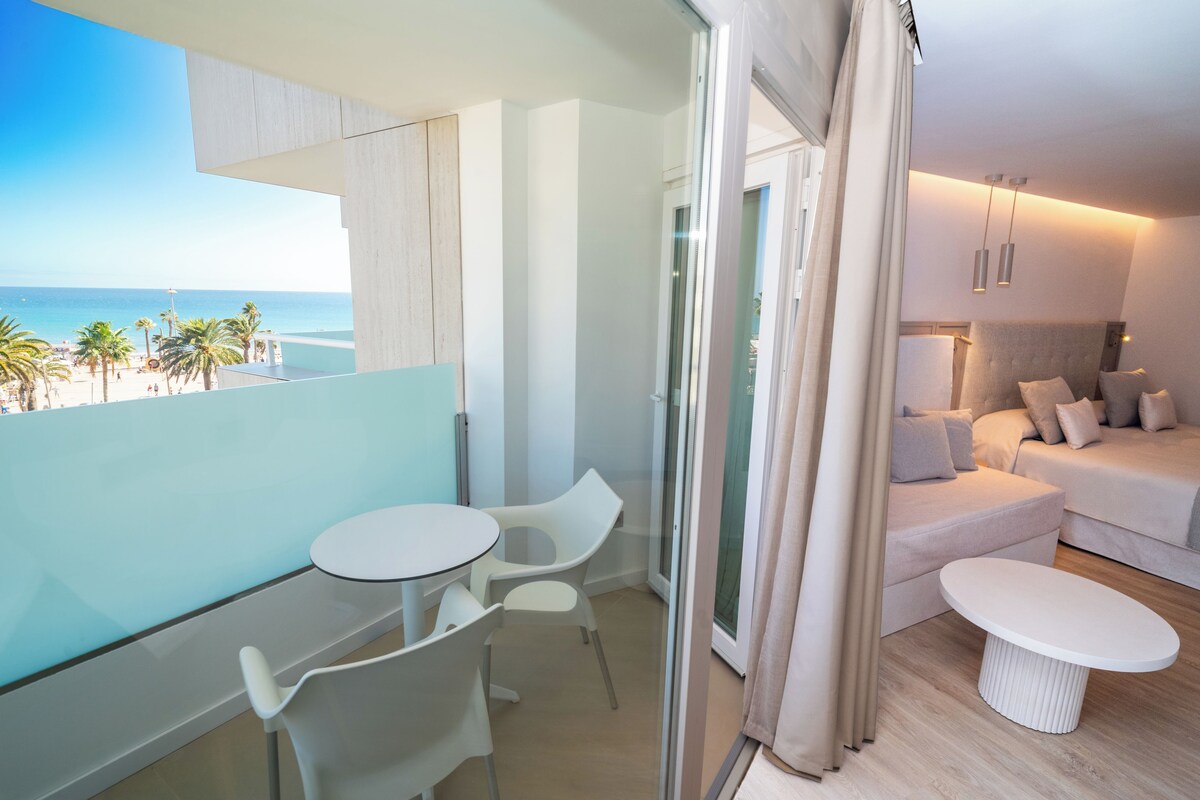 Varanda com vista para a praia, área de estar com mesa e cadeiras, e quarto com cama e sofá.