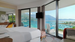 Quarto de hotel com uma cama grande, uma TV fixada na parede e uma varanda com vista para o mar e barcos.