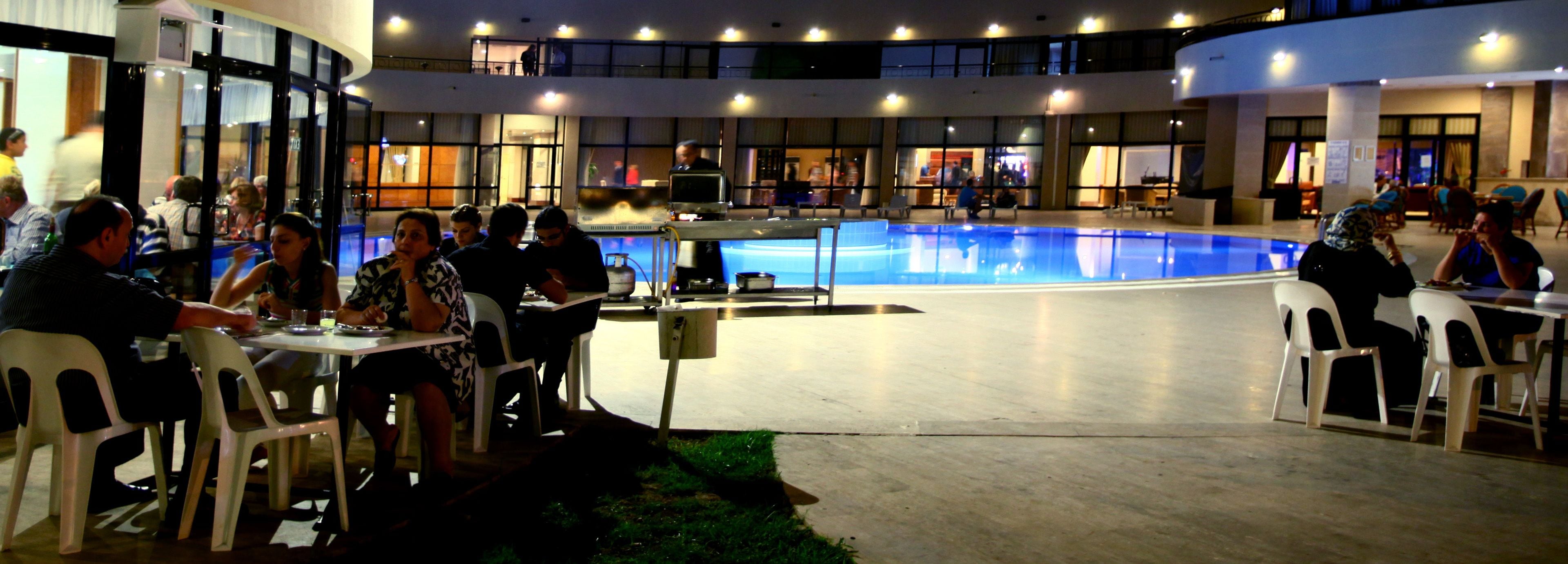 Área interna de piscina com mesas e cadeiras para refeições, ao redor de um edifício moderno com vários níveis e janelas amplas.