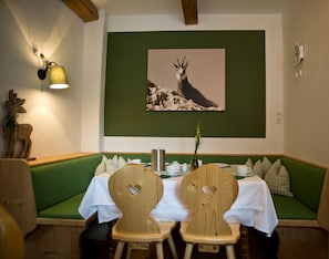 Uma área de refeições com um assento estofado verde, cadeiras de madeira, uma mesa posta com toalhas brancas, um quadro de um lhama e uma luminária fixada na parede.