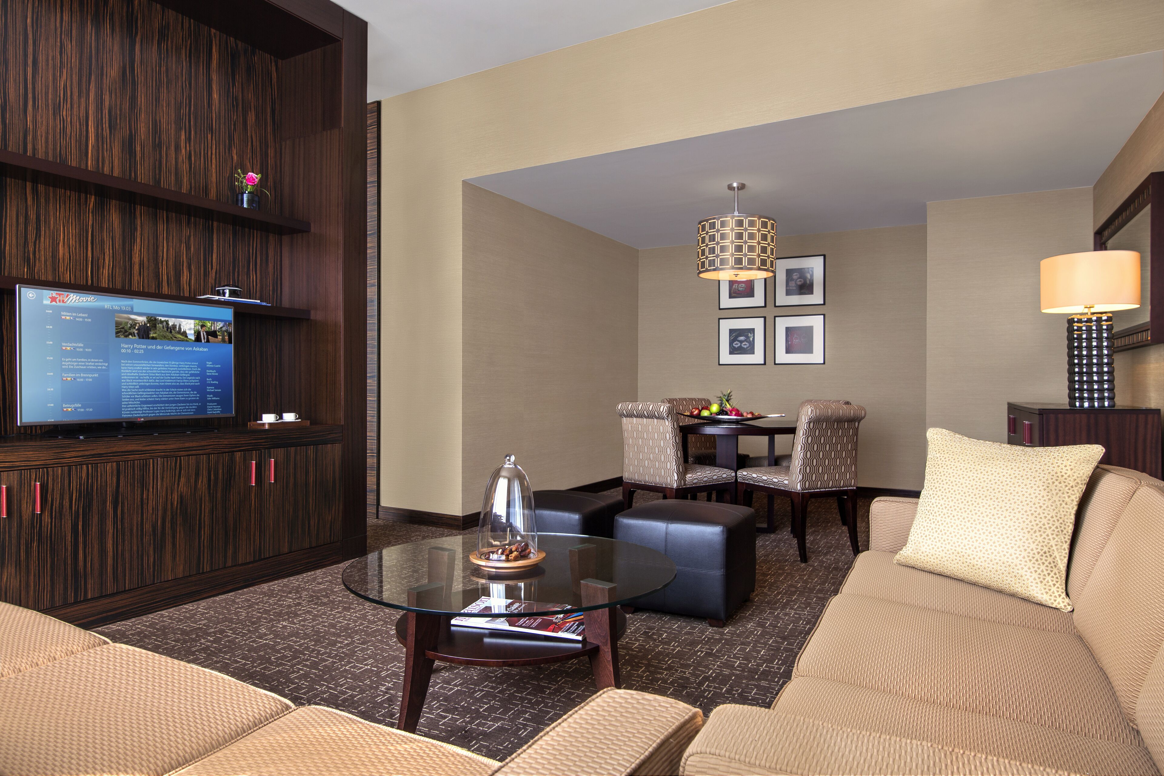 Quarto de hotel moderno com mesa de centro de vidro, sofá de couro e televisão de tela plana fixada em um armário de madeira.