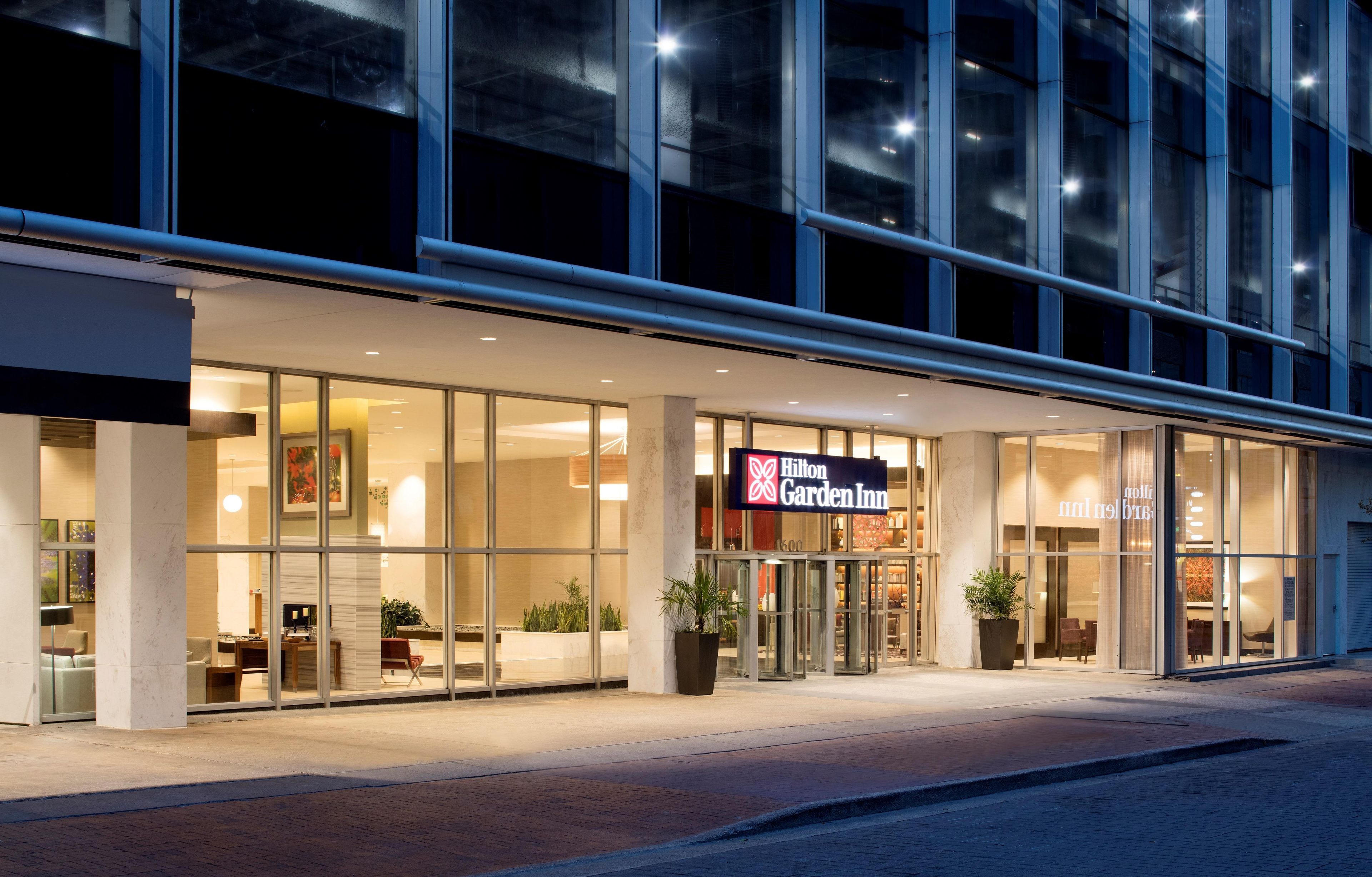 Entrada de um hotel com grandes portas de vidro, placa visível da Hilton Garden Inn e fachada de edifício moderna.