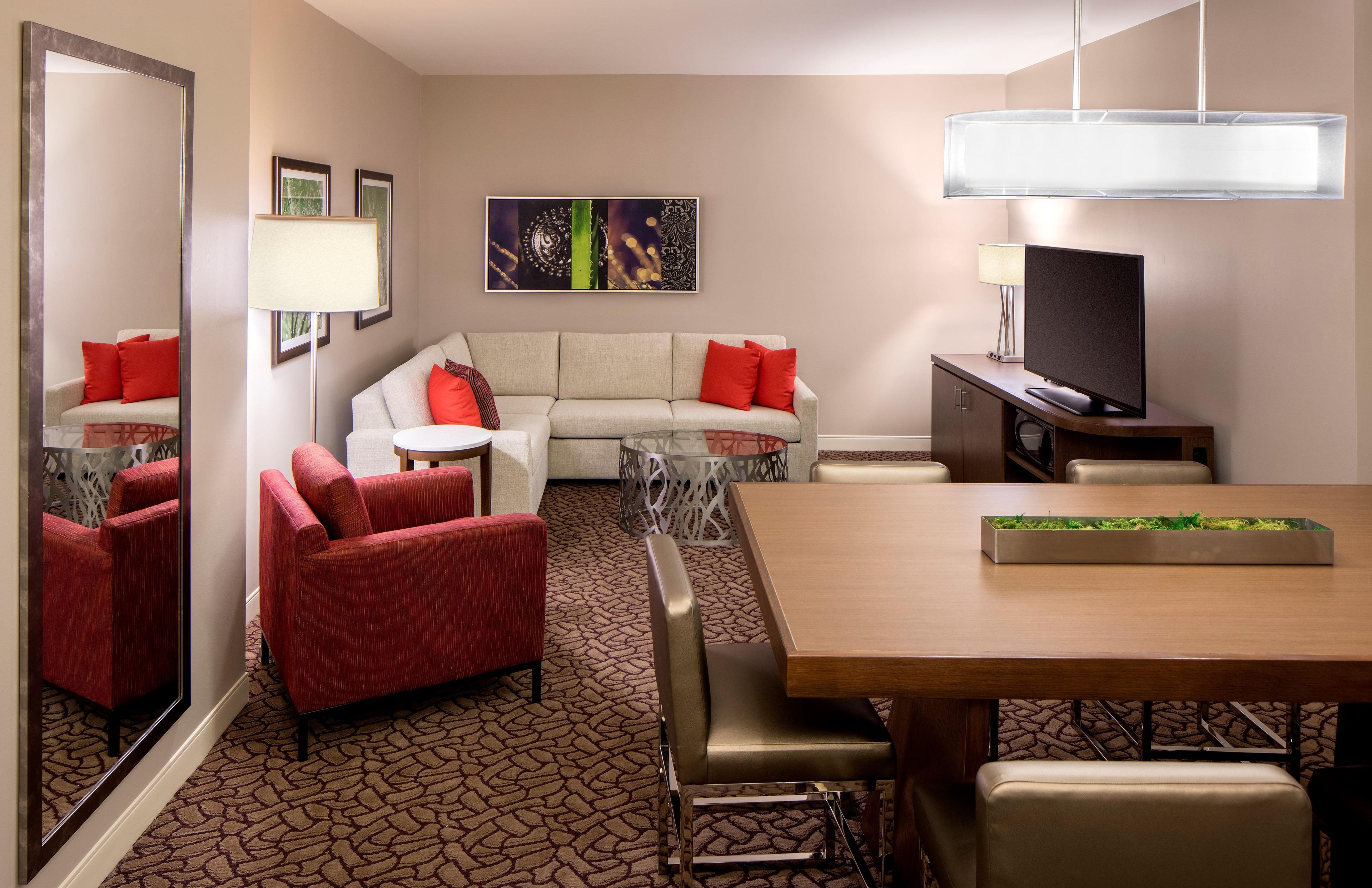 Quarto de hotel com sofá, poltronas, mesa de jantar, televisão e um objeto decorativo na parede.