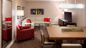 Quarto de hotel com sofá, poltronas, mesa de jantar, televisão e um objeto decorativo na parede.