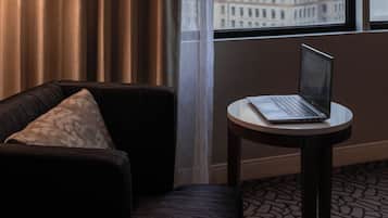 Quarto de hotel com vista para a paisagem urbana, um sofá com almofada estampada, uma mesa redonda com um laptop e uma janela grande com cortinas.