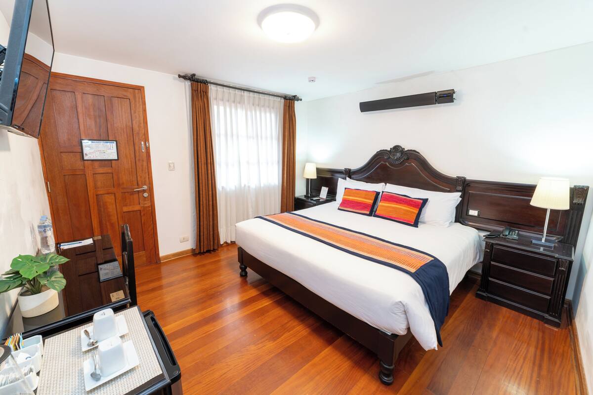 Quarto de hotel com piso de madeira, uma cama com cabeceira, mesa de cabeceira, escrivaninha com cadeira e janela com cortinas.