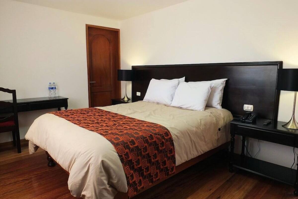 Quarto de hotel com cama, escrivaninha, cadeira, mesa de cabeceira, luminária e porta.