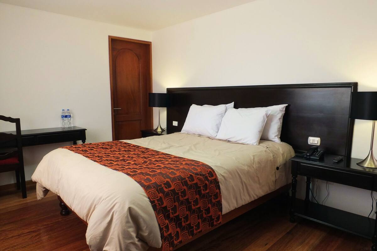 Quarto de hotel com cama, mesas de cabeceira, escrivaninha e porta.
