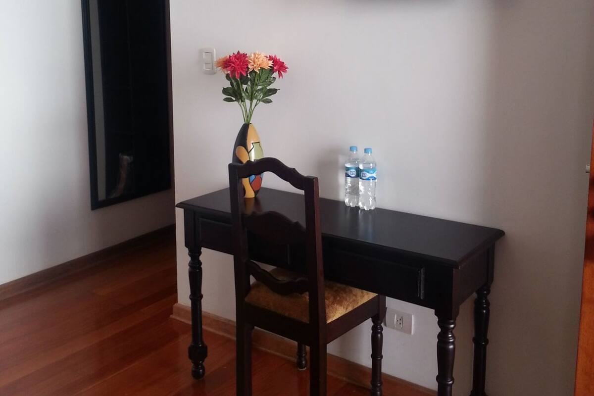 Um quarto com uma mesa de jantar de madeira, uma cadeira, um vaso com flores, uma garrafa de água e uma televisão de tela plana fixada na parede.
