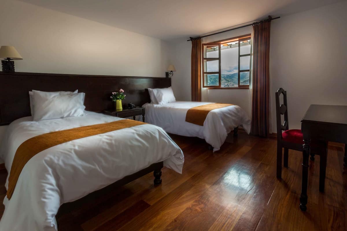 Quarto de hotel com duas camas, piso de madeira, janela com cortinas, mesa de cabeceira e uma cadeira.
