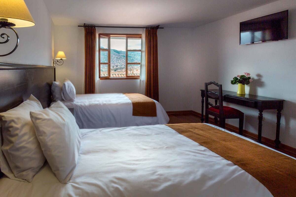 Quarto de hotel com duas camas, uma escrivaninha com cadeira, televisão na parede e janela com cortinas.
