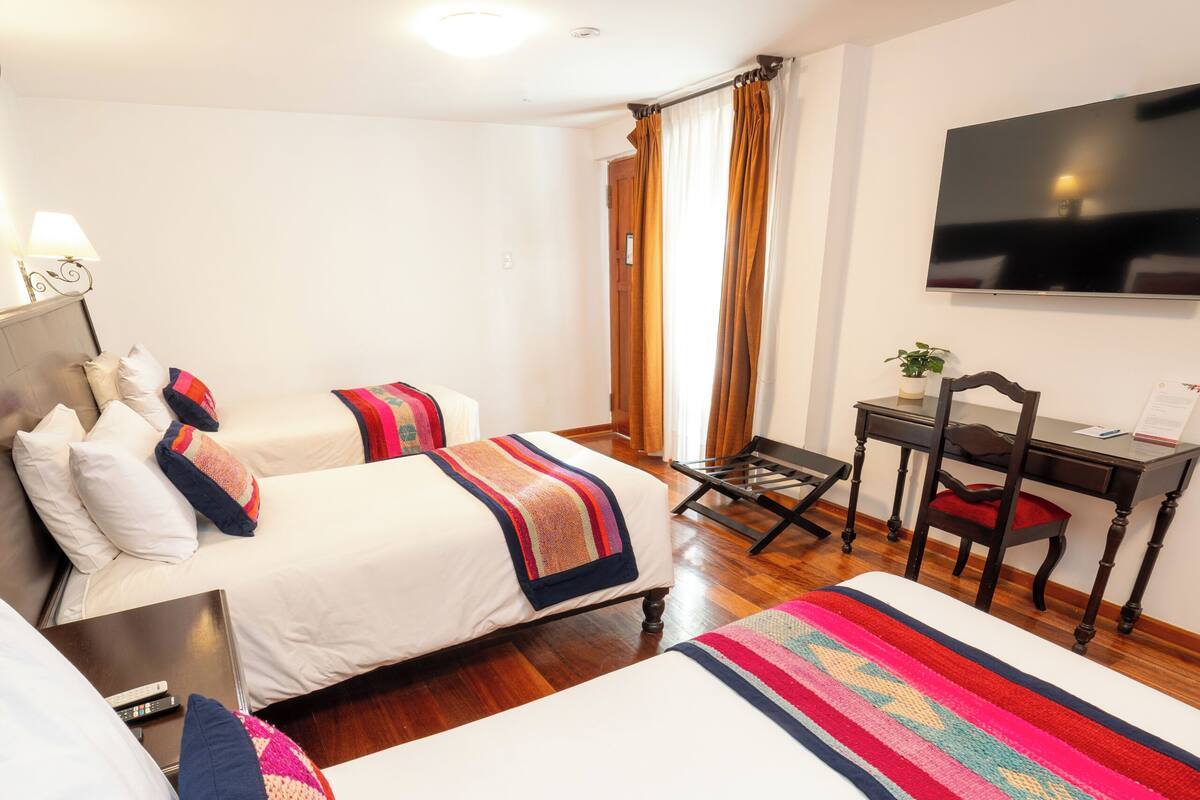 Quarto de hotel com duas camas, uma escrivaninha, uma cadeira, televisão e janela com cortinas.