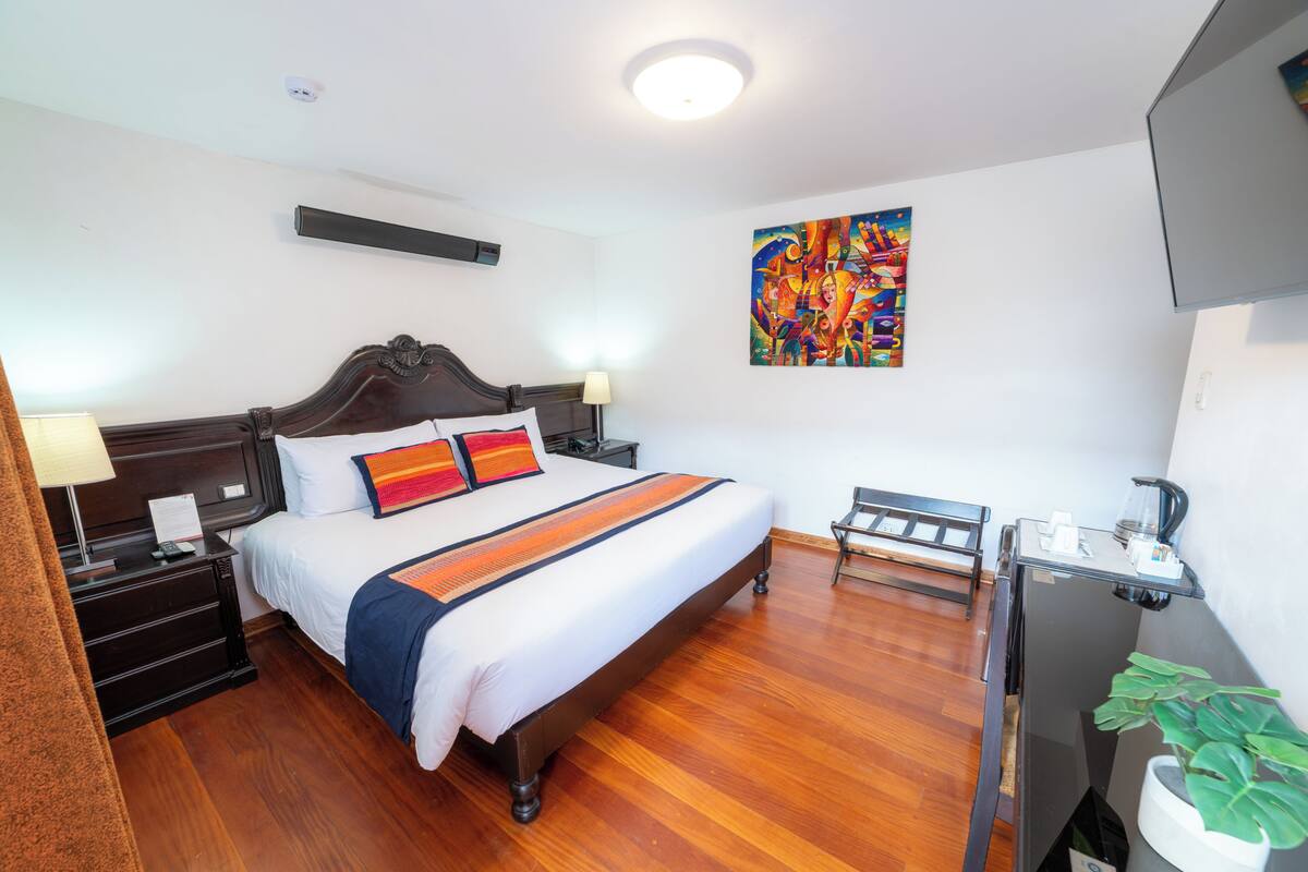 Quarto de hotel com cama de madeira, mesa de cabeceira, escrivaninha, cadeira, um quadro e uma planta.