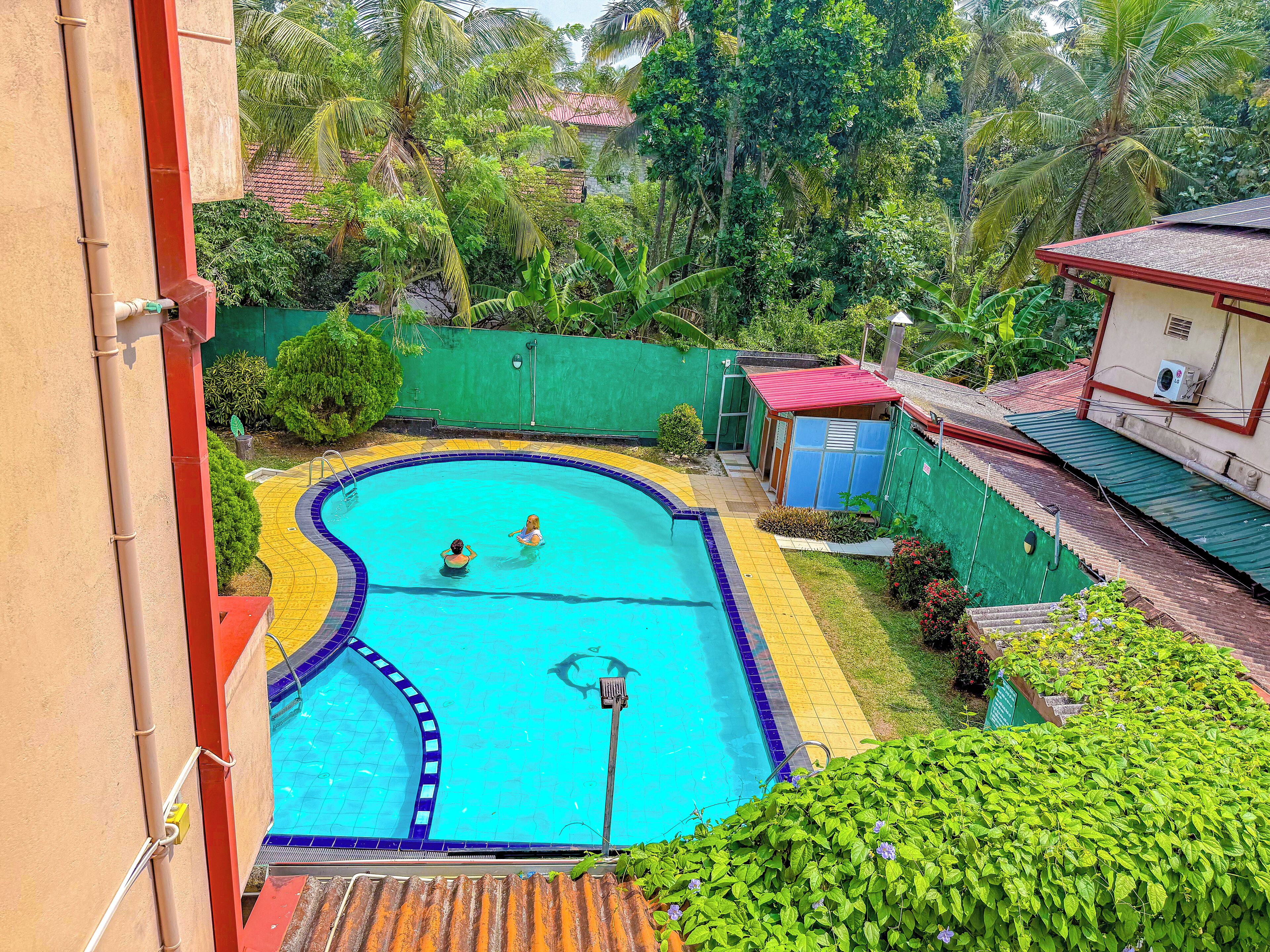 Piscina com duas pessoas na água, cercada por vegetação exuberante e uma pequena estrutura com telhado vermelho.