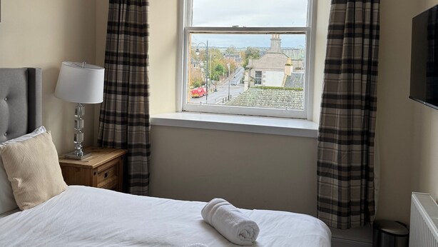 Quarto de hotel com uma cama, mesa de cabeceira com abajur, janela com cortinas e vista da cidade.