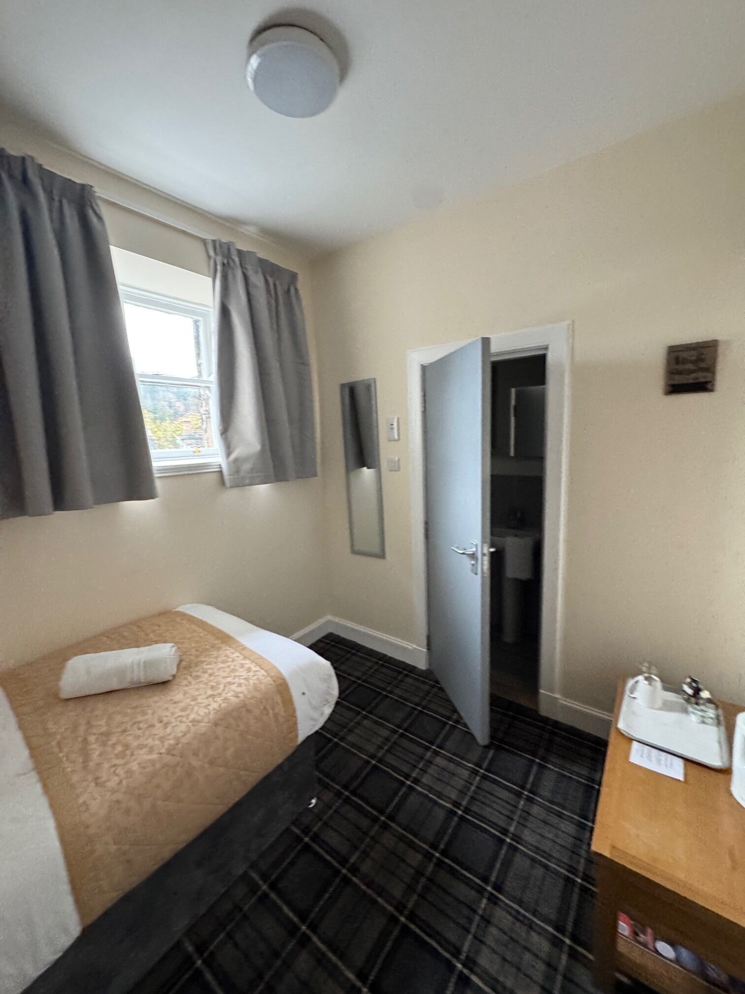 Quarto de hotel pequeno com uma cama de solteiro, mesa de cabeceira, espelho, janela com cortinas, porta e pia.