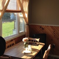 Uma mesa de jantar de madeira com um vaso de flores, saleiro e pimenteiro, e uma guardanapo. Uma janela com cortinas é visível, oferecendo vista para um gramado e um edifício.