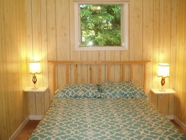 Um quarto com painéis de madeira, uma cama, duas mesas de cabeceira com abajures e uma janela com vista para árvores.