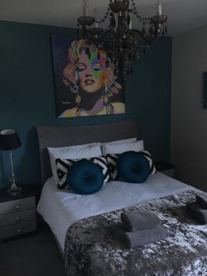 Quarto com cama, lustre, quadro na parede e mesa de cabeceira com abajur.