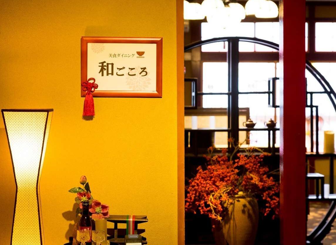 Quarto com parede amarela, um quadro com caracteres japoneses, um vaso com flores e uma janela com vista para um edifício.
