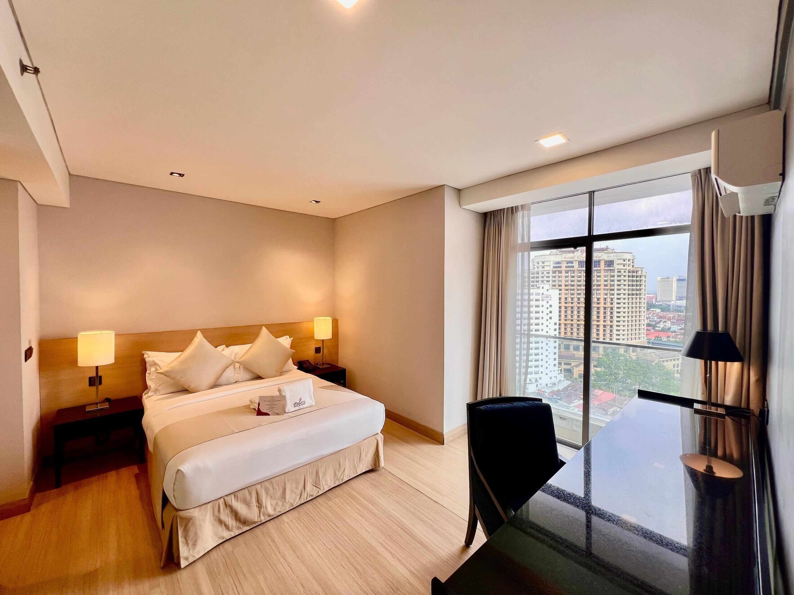 Quarto de hotel com uma cama grande, duas luminárias de cabeceira, uma cadeira e vista para a cidade através de uma janela ampla.