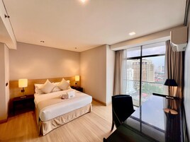 Quarto de hotel com uma cama grande, duas luminárias de cabeceira, uma cadeira e vista para a cidade através de uma janela ampla.