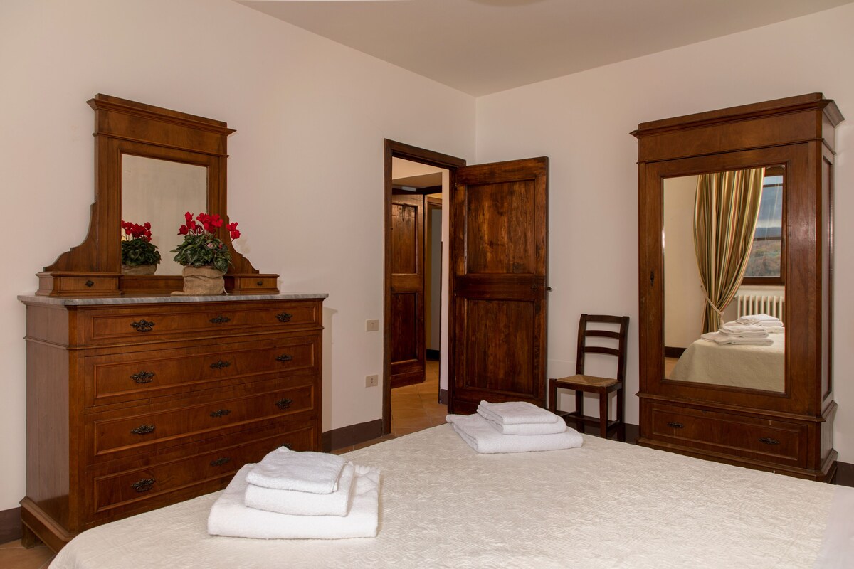 Quarto com cama grande, cómoda de madeira e espelho. Há toalhas na cama e uma cadeira no canto.