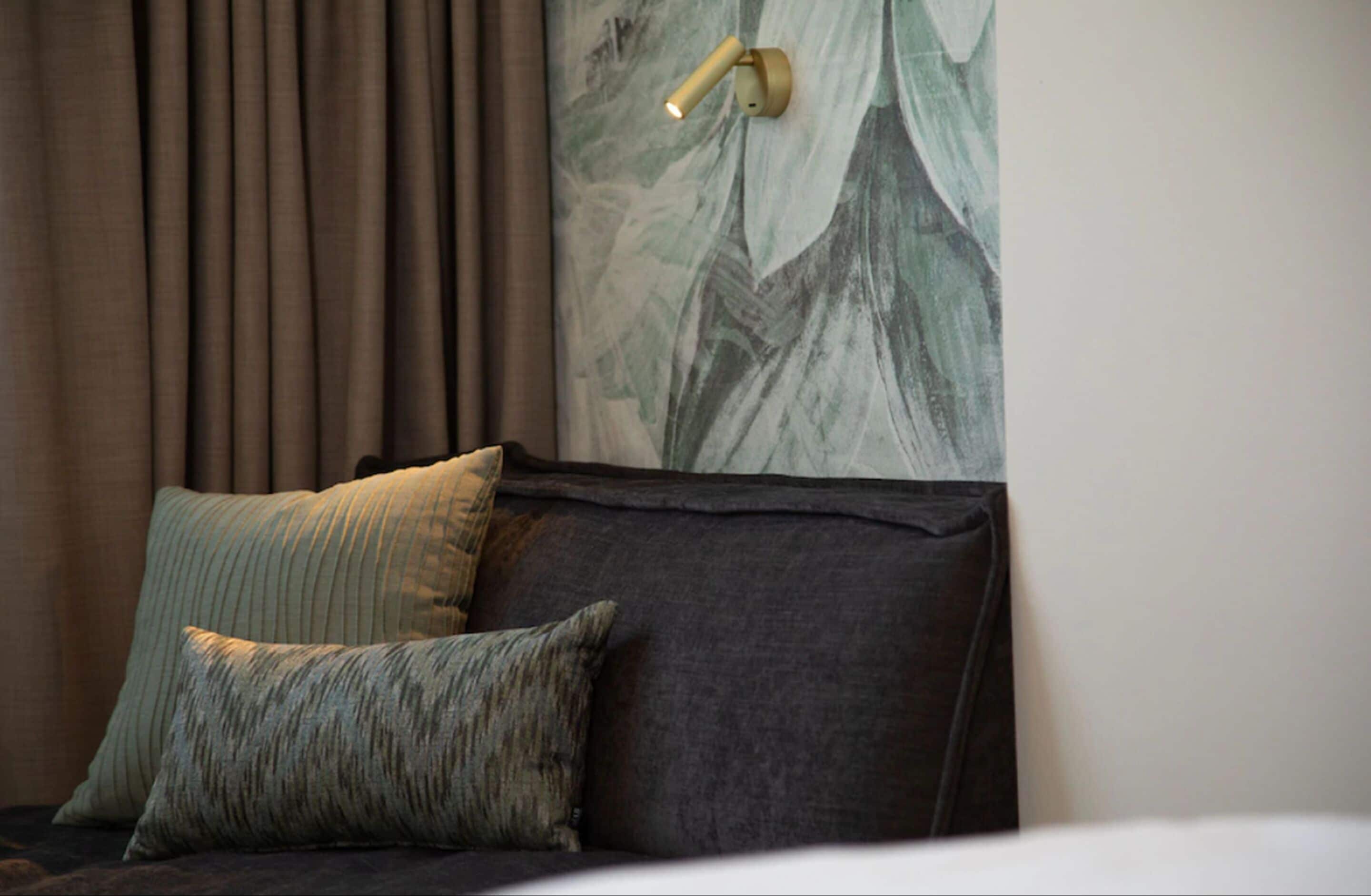 Uma cama com roupa de cama escura e duas almofadas, uma parede com pintura texturizada e uma luminária fixada na parede.