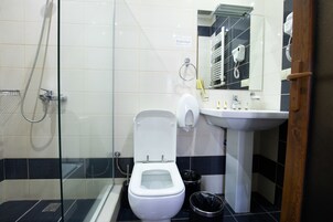 Uma casa de banho moderna com chuveiro em cabine de vidro, sanita branca, lavatório com espelho e um caixote do lixo preto.
