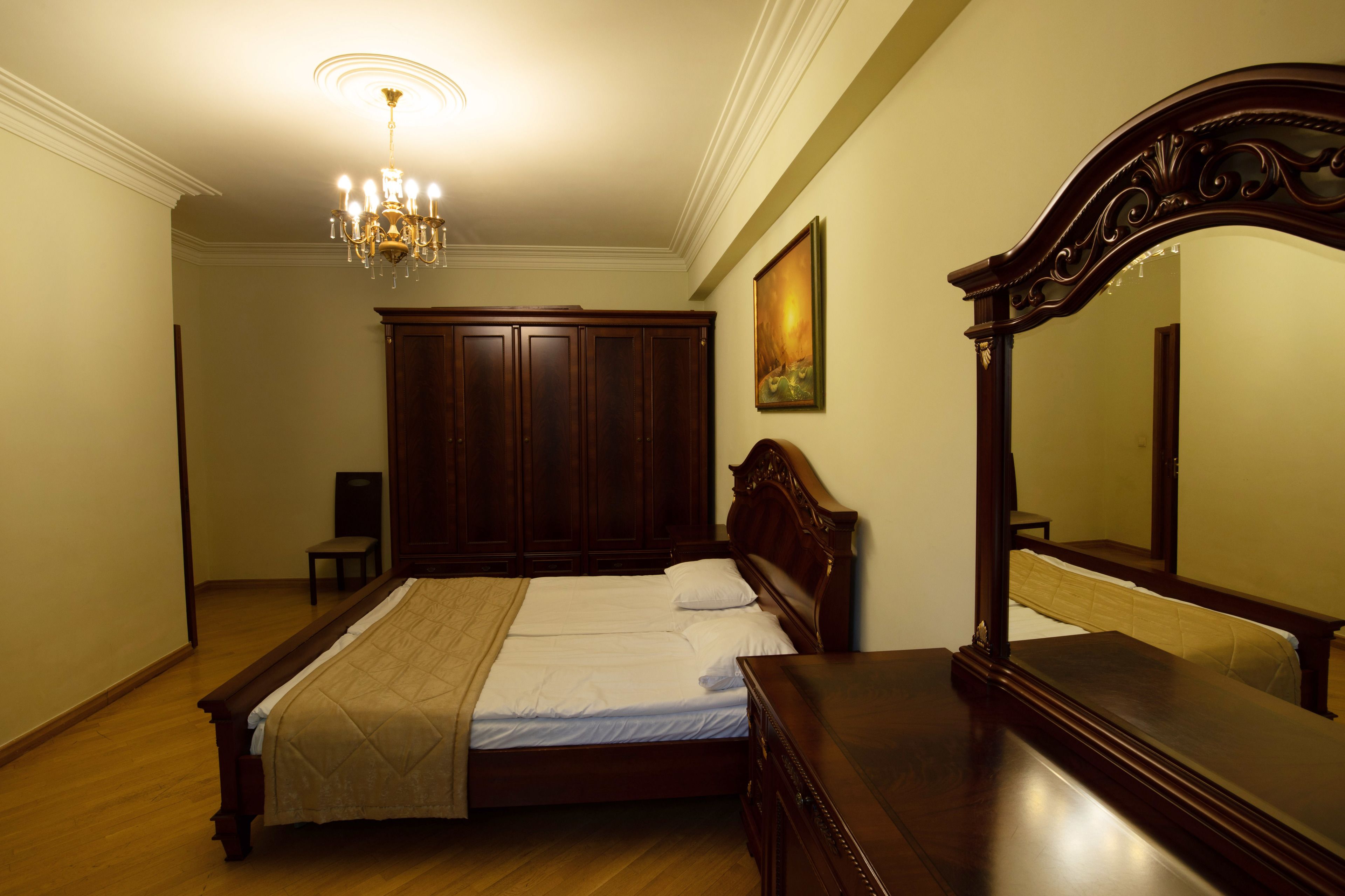 Um quarto com uma cama grande, cômoda de madeira, espelho, lustre e guarda-roupa.