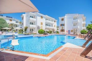 Piscina com deck revestido, cercada por edifícios brancos com varandas, e um céu azul límpido.
