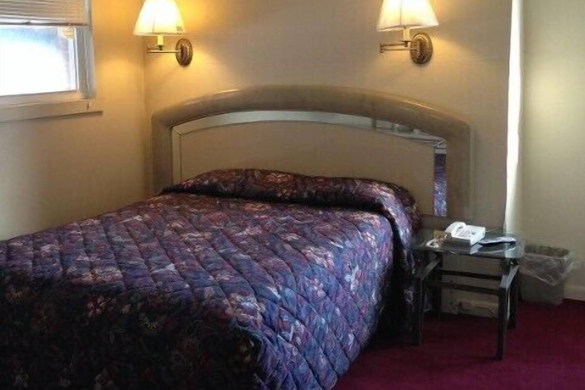 Quarto de hotel com uma cama, duas luminárias na parede, uma mesa de cabeceira e uma janela com persianas.